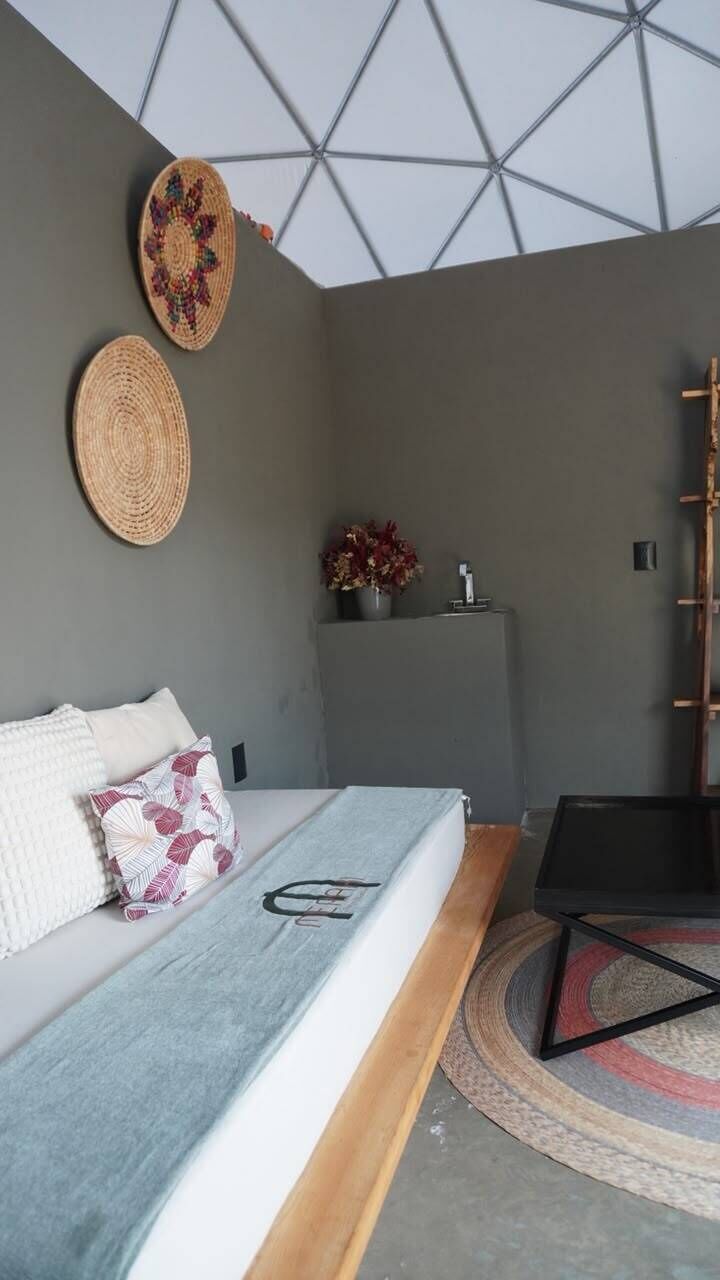 Quarto com uma cama, um cesto trançado fixado na parede, uma pequena mesa com um vaso e uma estrutura de teto geométrica.
