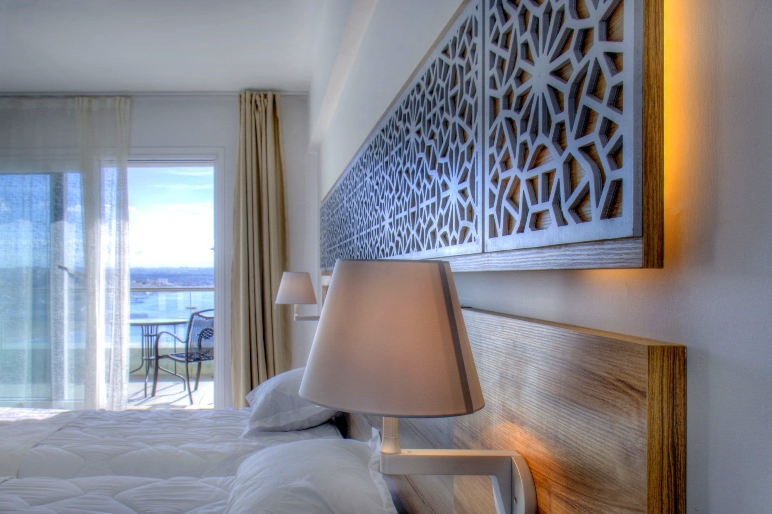 Quarto de hotel com cama, mesa de cabeceira com abajur, painel decorativo na parede e vista para o mar através de uma varanda.