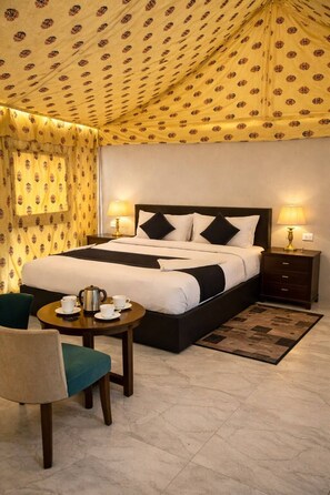 Quarto de hotel com uma cama grande, mesas de cabeceira com abajures, uma pequena mesa redonda com chávenas e um bule, e uma cadeira.