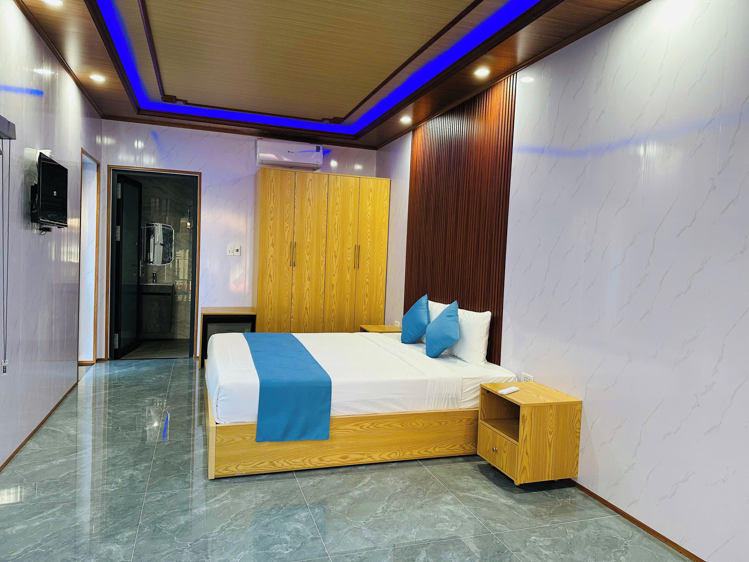 Quarto moderno com cama de madeira, mesa de cabeceira, guarda-roupa e banheiro visível através de uma porta aberta.