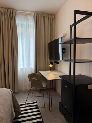 Quarto de hotel compacto com cama, escrivaninha, cadeira, televisão e guarda-roupa.