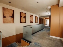 Uma área de recepção de hotel com design moderno, apresentando um balcão de recepção, uma escadaria e um corredor com obras de arte nas paredes.
