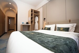 Quarto de hotel com uma cama grande, um roupão pendurado em um cabide, uma prateleira com amenities e uma porta que dá para outro quarto.