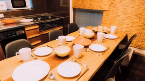 Uma mesa de jantar posta com pratos, tigelas, xícaras e talheres, rodeada por cadeiras em uma cozinha moderna com eletrodomésticos.