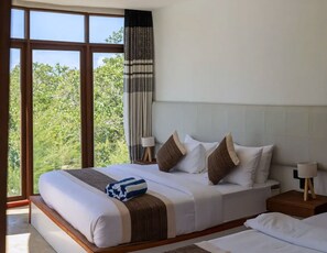 Quarto de hotel com uma cama grande, mesa de cabeceira com luminária e vista para o verde pela janela.