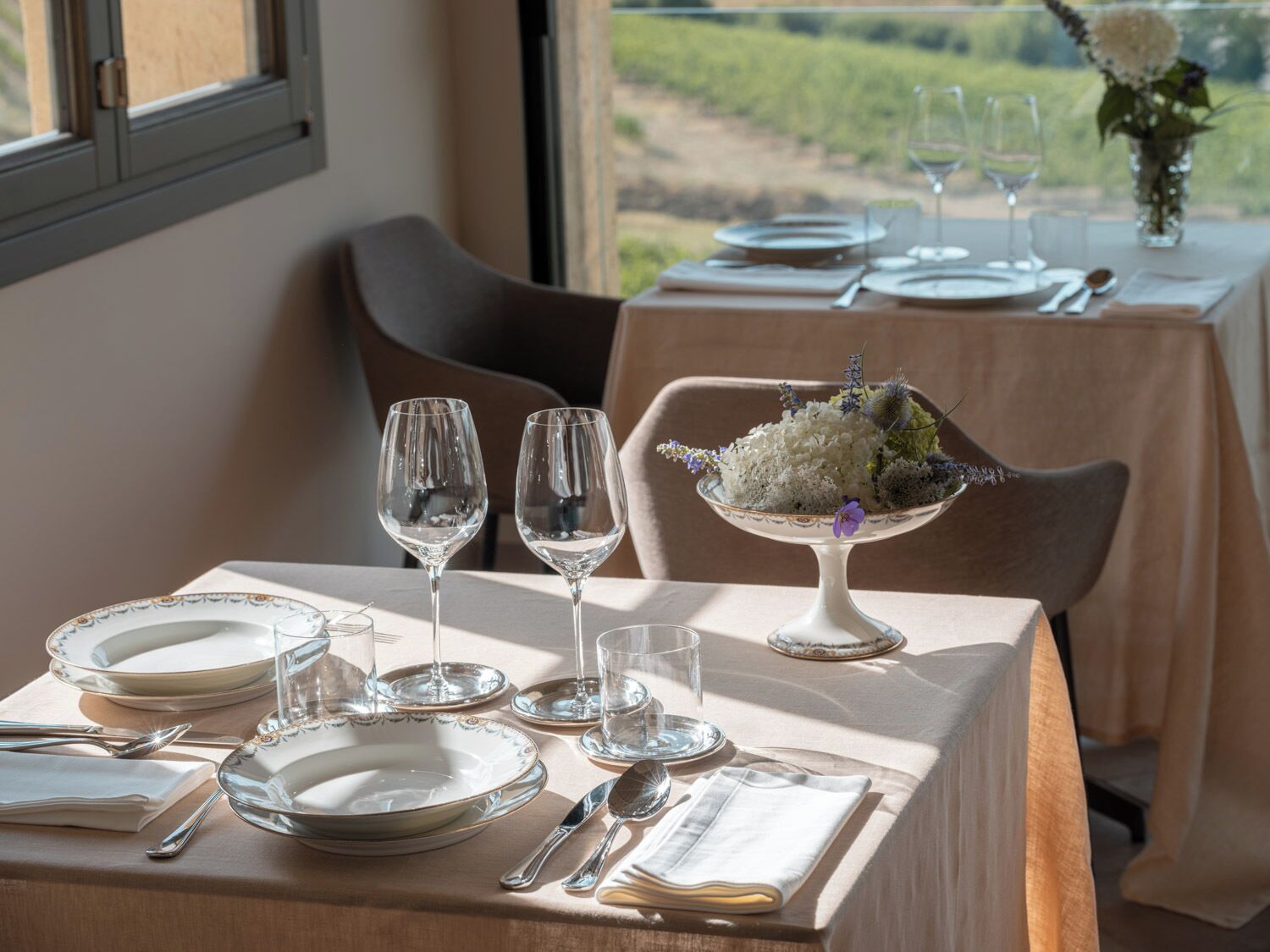 Mesa de jantar posta com pratos, copos e talheres, com um arranjo floral e vista para o exterior.