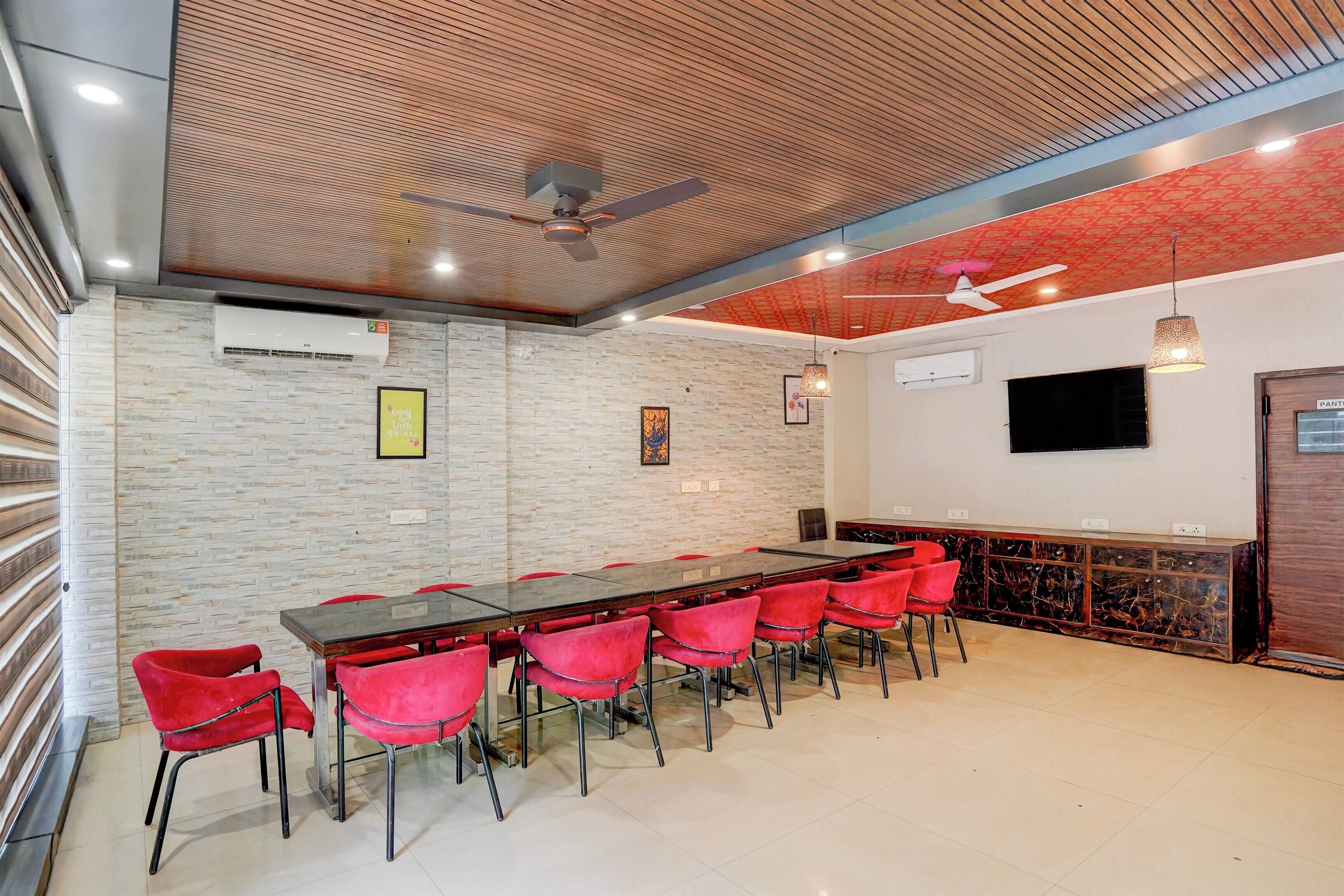Uma sala de conferências com uma mesa comprida, cadeiras vermelhas, um ventilador de teto e uma televisão instalada.