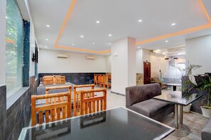 Um interior moderno com área de estar, incluindo cadeiras de madeira, um banco estofado e uma mesa com tampo de vidro. Cozinha visível através de um painel de vidro.