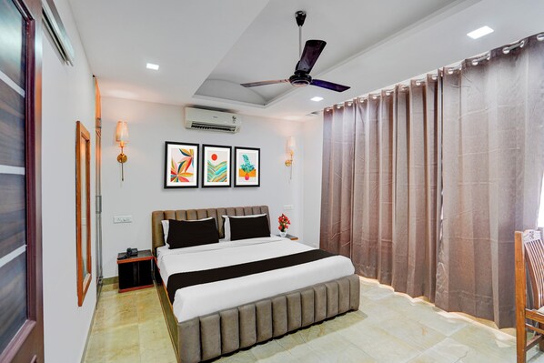 Quarto de hotel com cama, ventoinha no teto, aparelho de ar condicionado, mesa de cabeceira, luminária e janela com cortinas.
