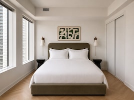 Quarto moderno com uma cama grande, duas mesas de cabeceira, uma obra de arte na parede e vista para um edifício pela janela.