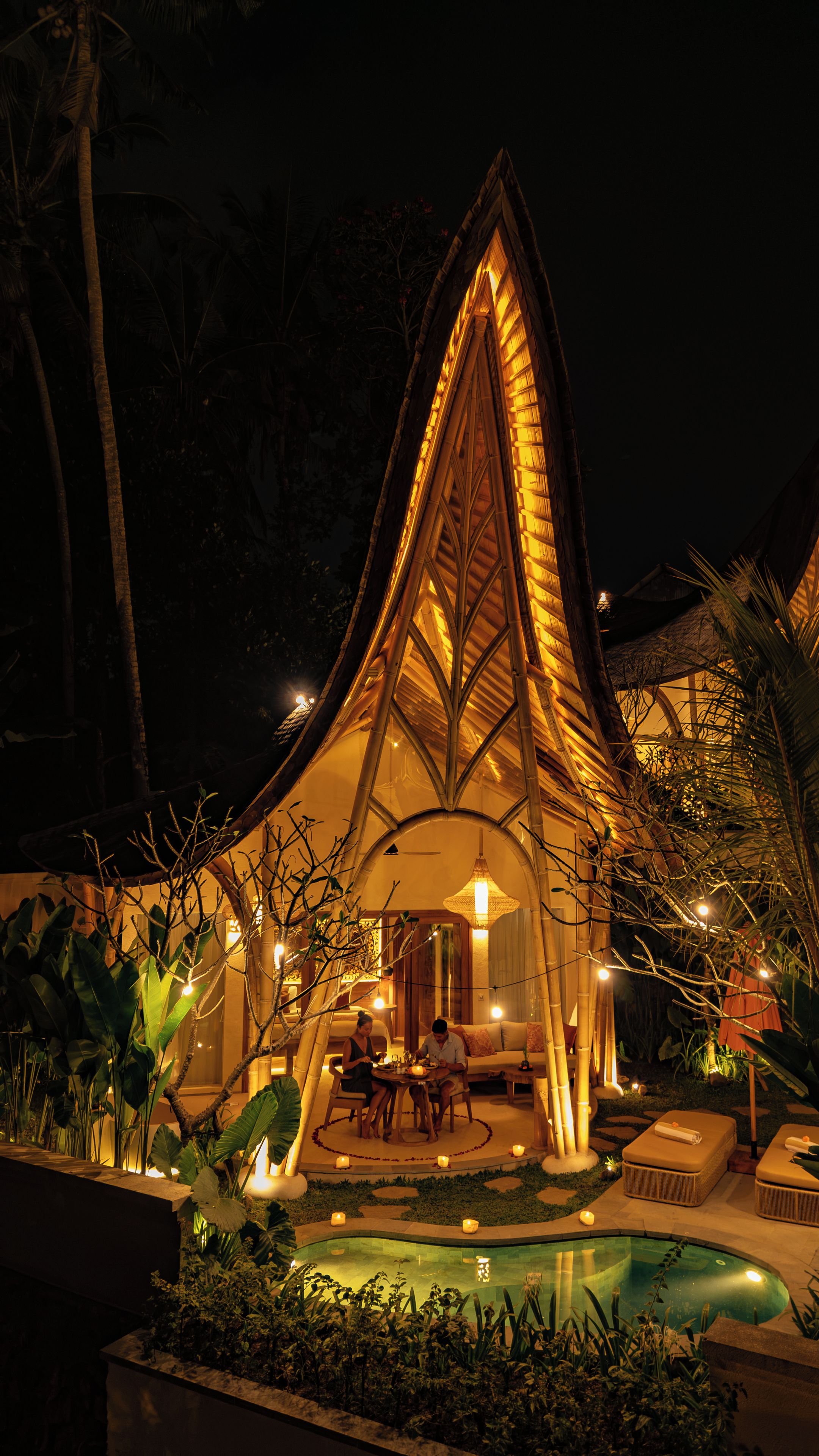 Um edifício triangular com fachada de vidro, cercado por vegetação exuberante e uma piscina.