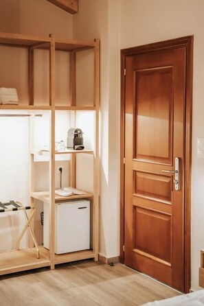 Quarto com porta de madeira, uma pequena geladeira branca e uma estante de madeira com chaleira e xícaras.