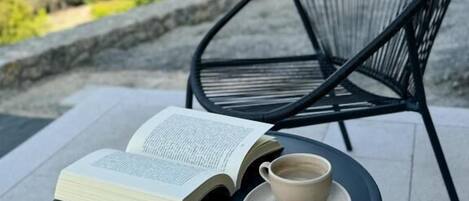 Um livro aberto sobre uma mesa, com uma xícara e um biscoito, em ambiente externo, com uma cadeira e vista para árvores e colinas.