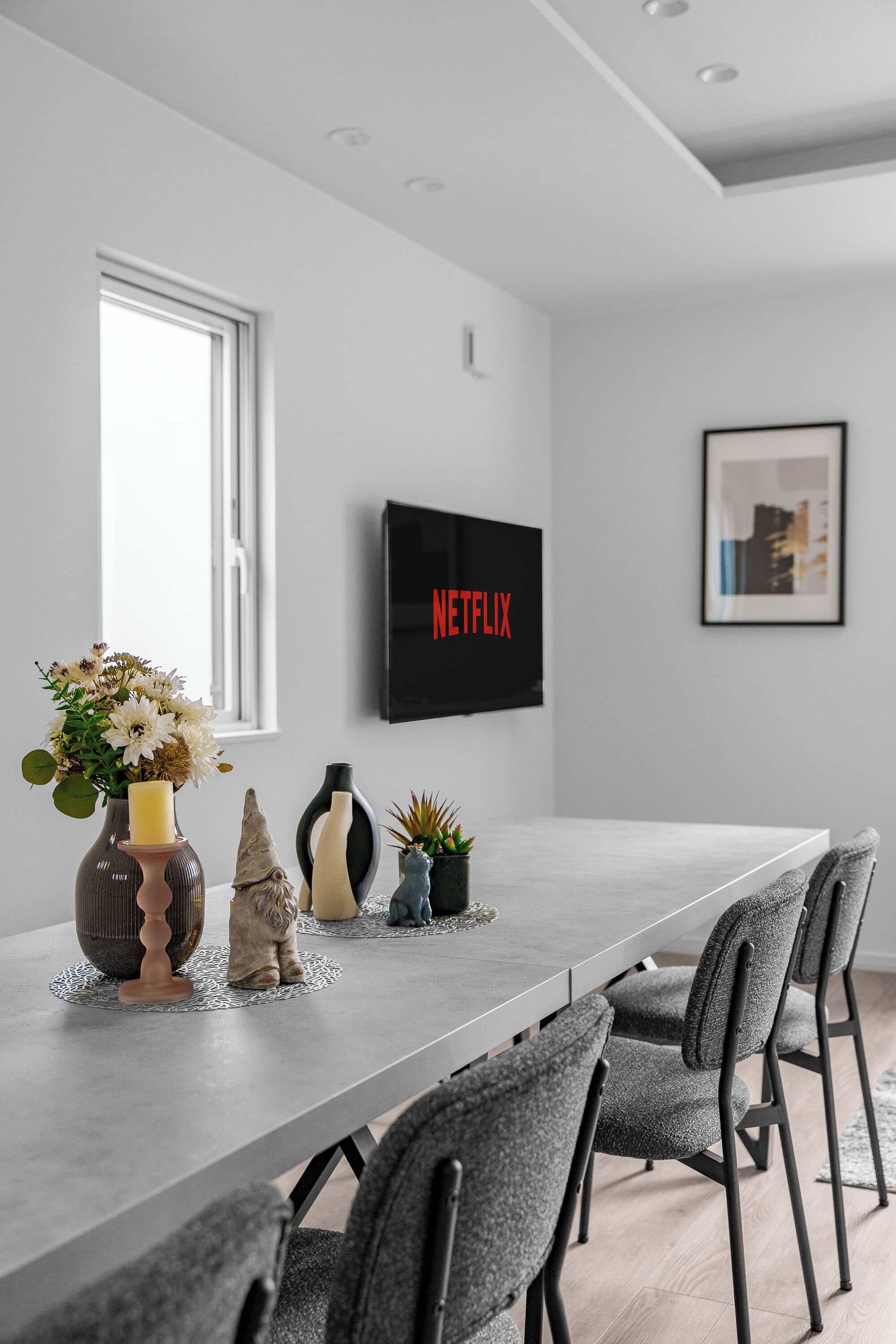 Uma área de jantar moderna com uma mesa comprida, cadeiras, um vaso com flores, uma vela e uma TV fixada na parede exibindo o logotipo da Netflix.