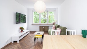 Uma sala de estar moderna com uma janela grande, um sofá branco, uma mesa de jantar e uma televisão fixada na parede.