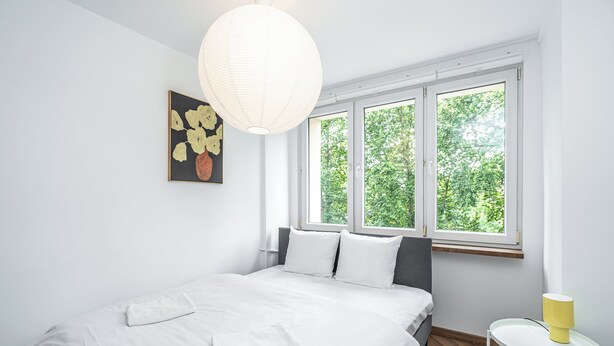 Quarto com cama, mesa de cabeceira, luminária e janela com vista para área verde.