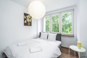 Quarto com cama, mesa de cabeceira, luminária e janela com vista para área verde.