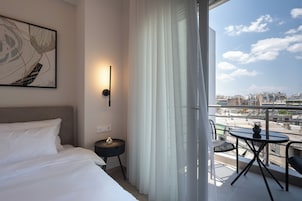 Quarto de hotel com cama, mesa de cabeceira com um vaso, luminária na parede, cortinas finas e varanda com mesa e cadeiras.
