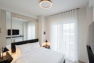 Quarto de hotel com cama, mesa de cabeceira, luminária, televisão, espelho grande e cortinas translúcidas na janela.