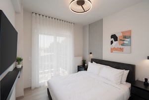 Quarto moderno com uma cama grande, uma televisão de tela plana, uma mesa de cabeceira, um vaso de planta e uma janela com cortinas translúcidas.
