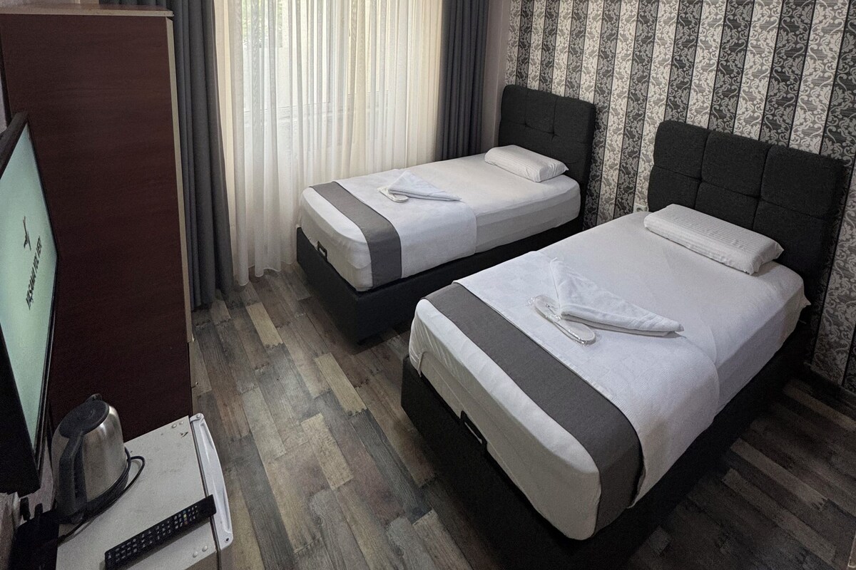 Quarto de hotel com duas camas, uma mesa de escritório com um computador e uma chaleira, e uma janela com cortinas.