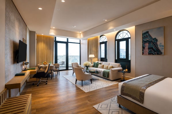 Quarto de hotel moderno com cama, sofá, escrivaninha com cadeira, área de refeições e grandes janelas.