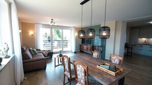 Uma sala de jantar ampla com mesa e cadeiras de madeira, uma cozinha ao fundo e uma grande janela que proporciona luz natural.