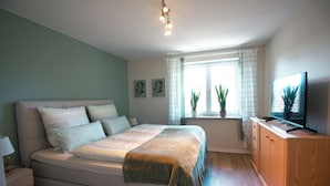 Quarto com cama, mesa de cabeceira, luminária, janela com cortinas, plantas em vasos e uma televisão sobre um armário de madeira.