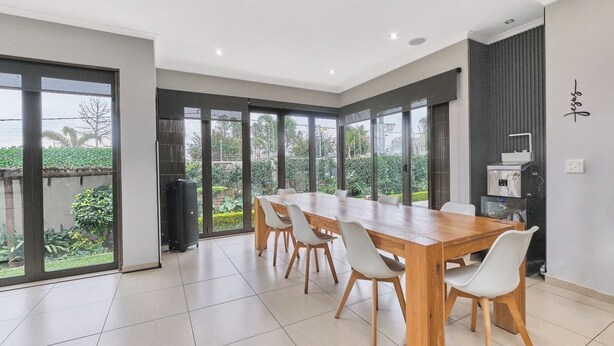 Uma sala de jantar moderna com uma longa mesa de madeira e cadeiras brancas, grandes janelas com vista para o verde, e uma área de cozinha com eletrodomésticos.