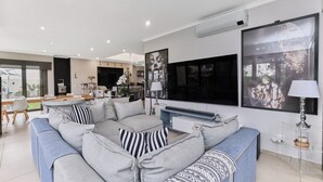 Uma sala de estar moderna com um sofá cinzento, uma televisão de tela plana e um espaço de jantar com mesa e cadeiras.