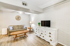 Uma sala de estar com parede de tijolos brancos, um sofá bege, uma mesa de centro de madeira, um armário branco com gavetas, uma televisão e uma pequena mesa com um abajur e planta.