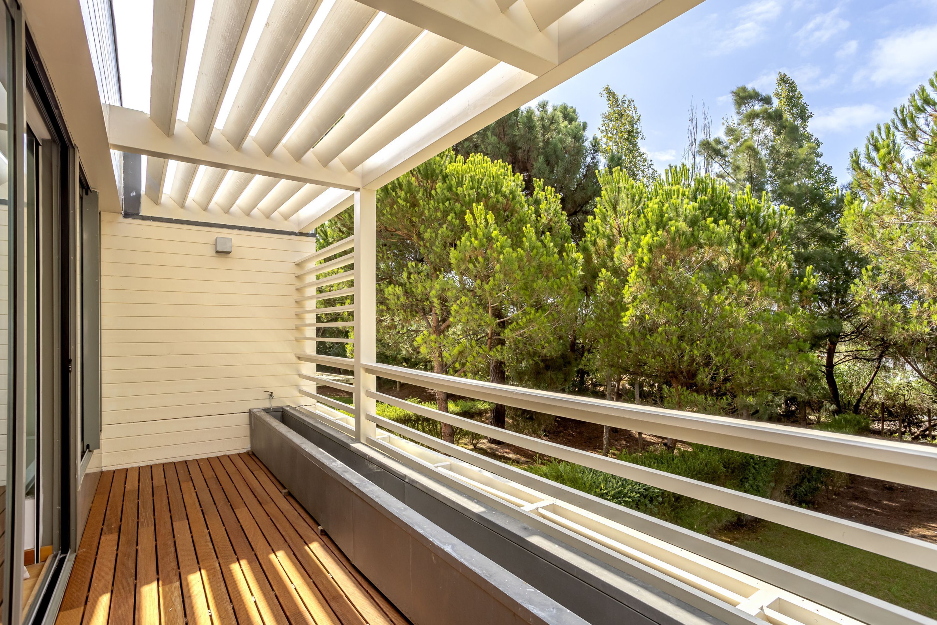 Varanda com piso de madeira e guarda-corpo branco, com vista para um jardim de pinheiros.
