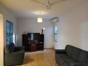 Uma sala de estar com televisão de tela plana, dois sofás, uma mesa de centro, ventilador de teto e uma janela com persianas.