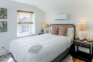 Uma cama bem arrumada com roupa de cama branca e uma toalha dobrada, uma mesa de cabeceira com abajur, um ar condicionado fixado na parede e uma janela com persianas.