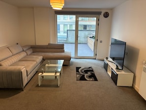 Uma sala de estar moderna com um sofá, uma mesa de centro de vidro, uma TV de tela plana e uma porta de correr de vidro que dá para uma varanda.
