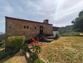 Um edifício de pedra com telhado de telhas vermelhas, um pequeno alpendre e um jardim com flores vermelhas.