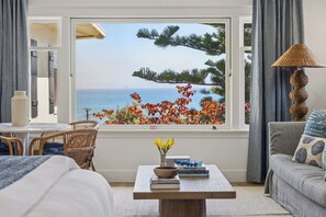 Quarto com vista para o mar, janela com vista para árvores e flores, mesa com livros e um vaso, e um abajur.