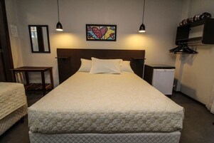 Quarto de hotel com uma cama grande, mesas de cabeceira, abajur, pequena escrivaninha, uma cadeira e televisão na parede.