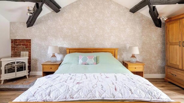 Quarto com teto inclinado, cama com colcha floral, duas luminárias de cabeceira, guarda-roupa de madeira e um armário branco com porta de vidro.
