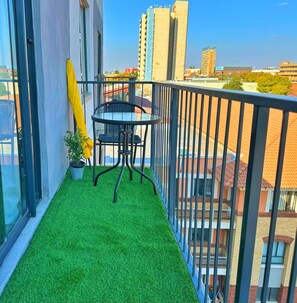 Varanda com grama sintética, uma mesa redonda pequena e cadeiras, com vista para um horizonte urbano com edifícios.