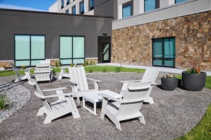 Uma área externa com assentos, incluindo cadeiras Adirondack brancas e mesas, um muro de pedra e um edifício com grandes janelas.