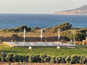 Uma área na piscina com espreguiçadeiras e guarda-sóis, um muro de pedra e vista para o mar e falésias.