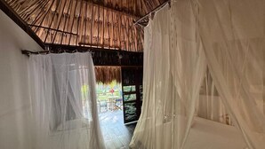 Quarto com telhado de colmo, cama com dossel e vista para área externa com mesas e cadeiras.