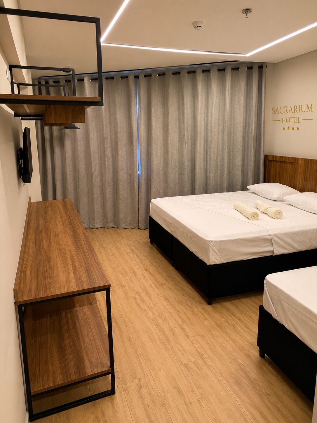 Quarto de hotel com cama, escrivaninha e televisão fixada na parede.