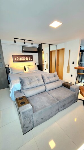 Quarto moderno com um amplo sofá cinzento, uma cama com cabeceira e uma obra de arte na parede retratando as fases da lua.