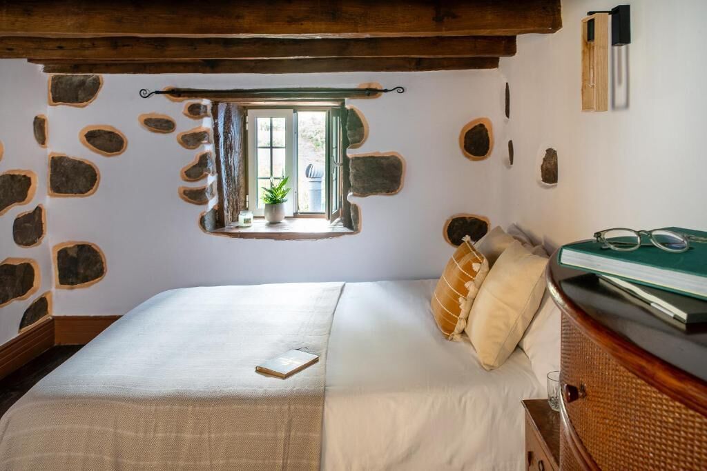 Quarto com parede de pedra, uma janela, cama com almofadas, mesa de cabeceira e um livro.