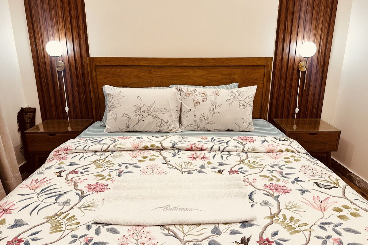 Cama bem arrumada com roupa de cama floral, duas luminárias de cabeceira e cabeceira de madeira.