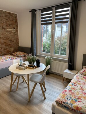 Quarto aconchegante com parede de tijolos, janela com persianas, mesa redonda com cadeira, cama com roupa de cama floral e radiador.