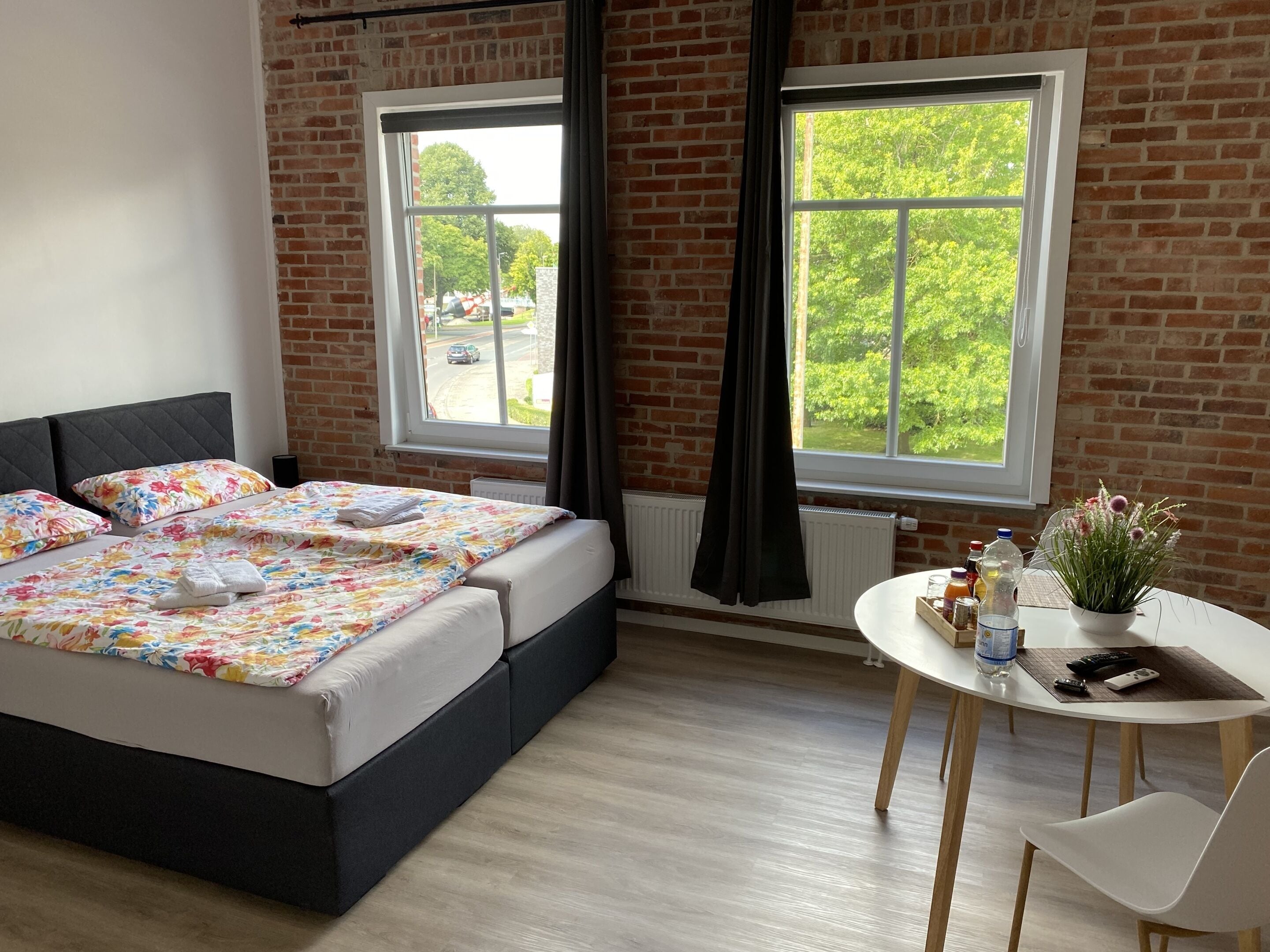 Quarto de hotel com cama, mesa com planta e vista para o exterior através de grandes janelas.
