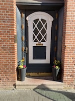 Uma porta preta com janela em padrão de losangos e placa de latão, ladeada por paredes de tijolo e dois vasos de flores.